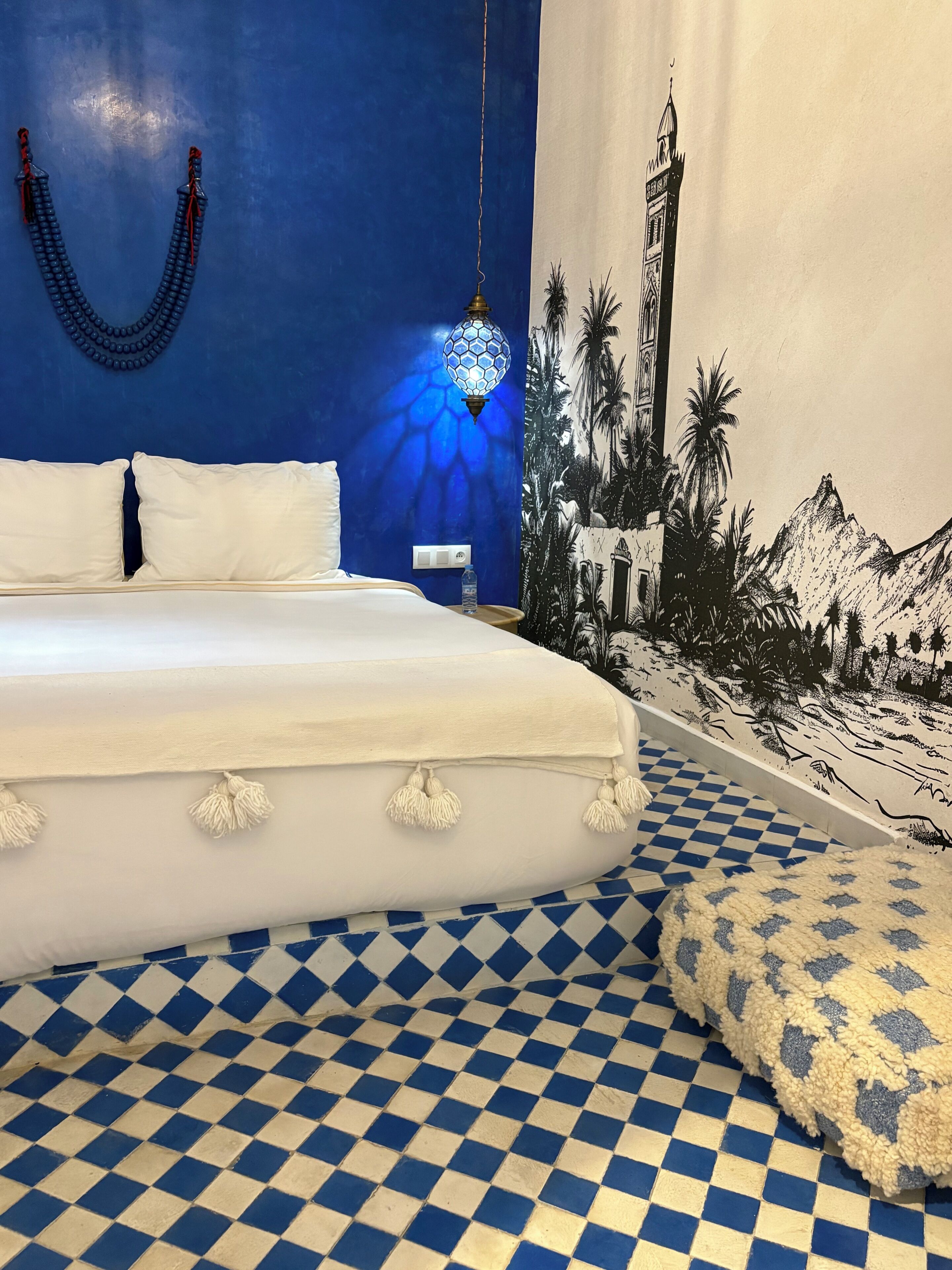 Uma cama com roupa de cama branca, um chão quadriculado azul e branco, uma luminária fixa na parede e um mural com paisagem de montanhas.