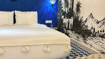 Uma cama com roupa de cama branca, um chão quadriculado azul e branco, uma luminária fixa na parede e um mural com paisagem de montanhas.