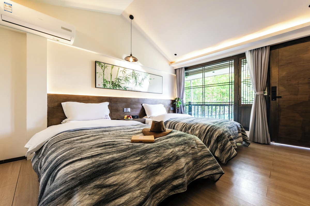 Quarto de hotel com duas camas, uma janela grande com vista para o verde, ar-condicionado na parede e uma luminária pendente.