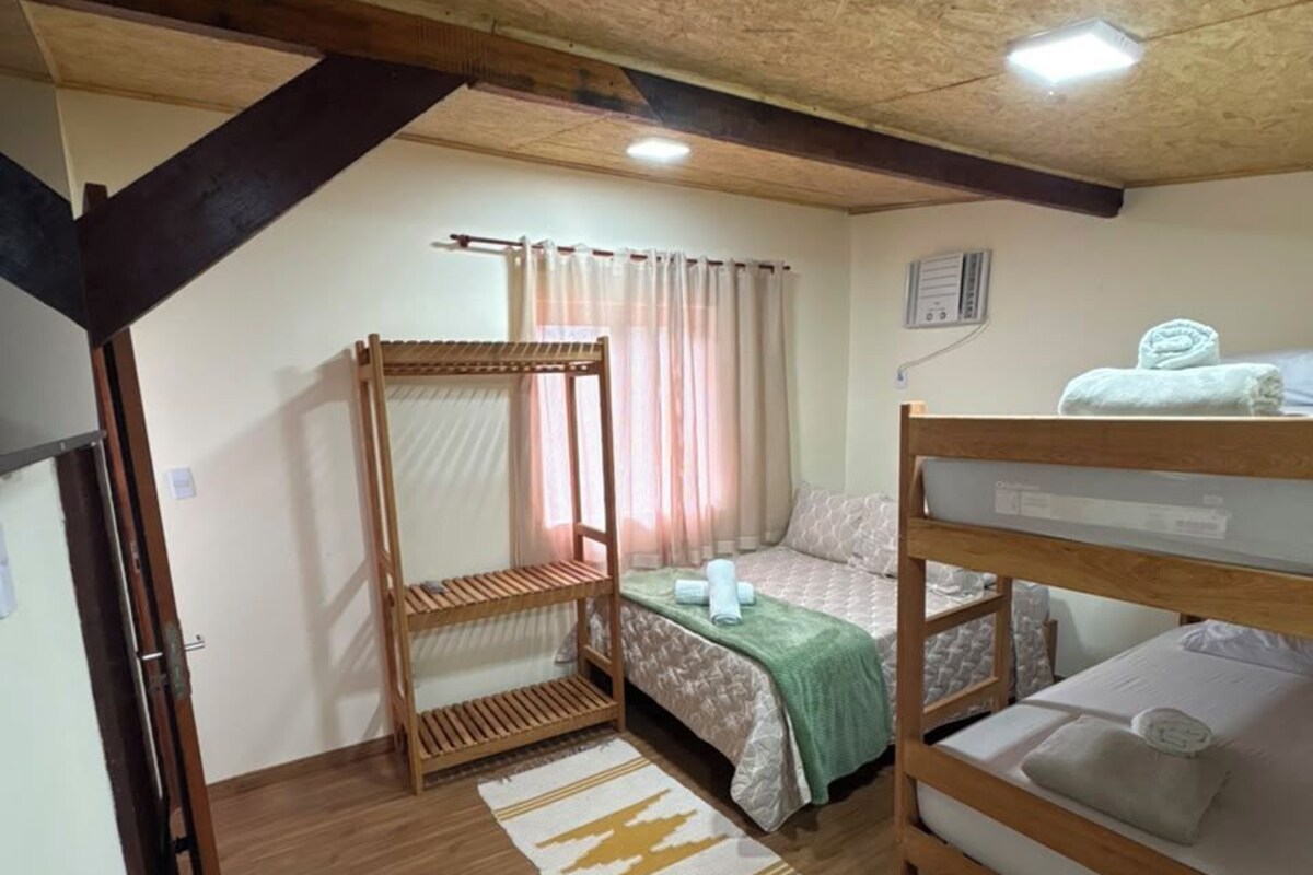 Quarto pequeno e acolhedor com beliche, cama de solteiro, teto de madeira e janela com cortinas.