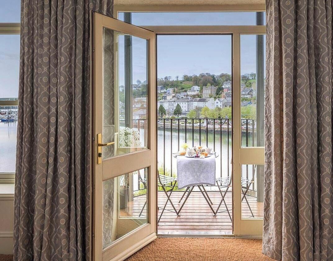Quarto com porta de vidro que dá para uma varanda externa com mesa e cadeiras, com vista para um corpo d'água e edifícios.
