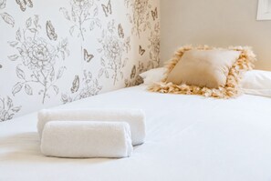 Uma cama com roupa de cama branca, um travesseiro fofo e um padrão de papel de parede floral.