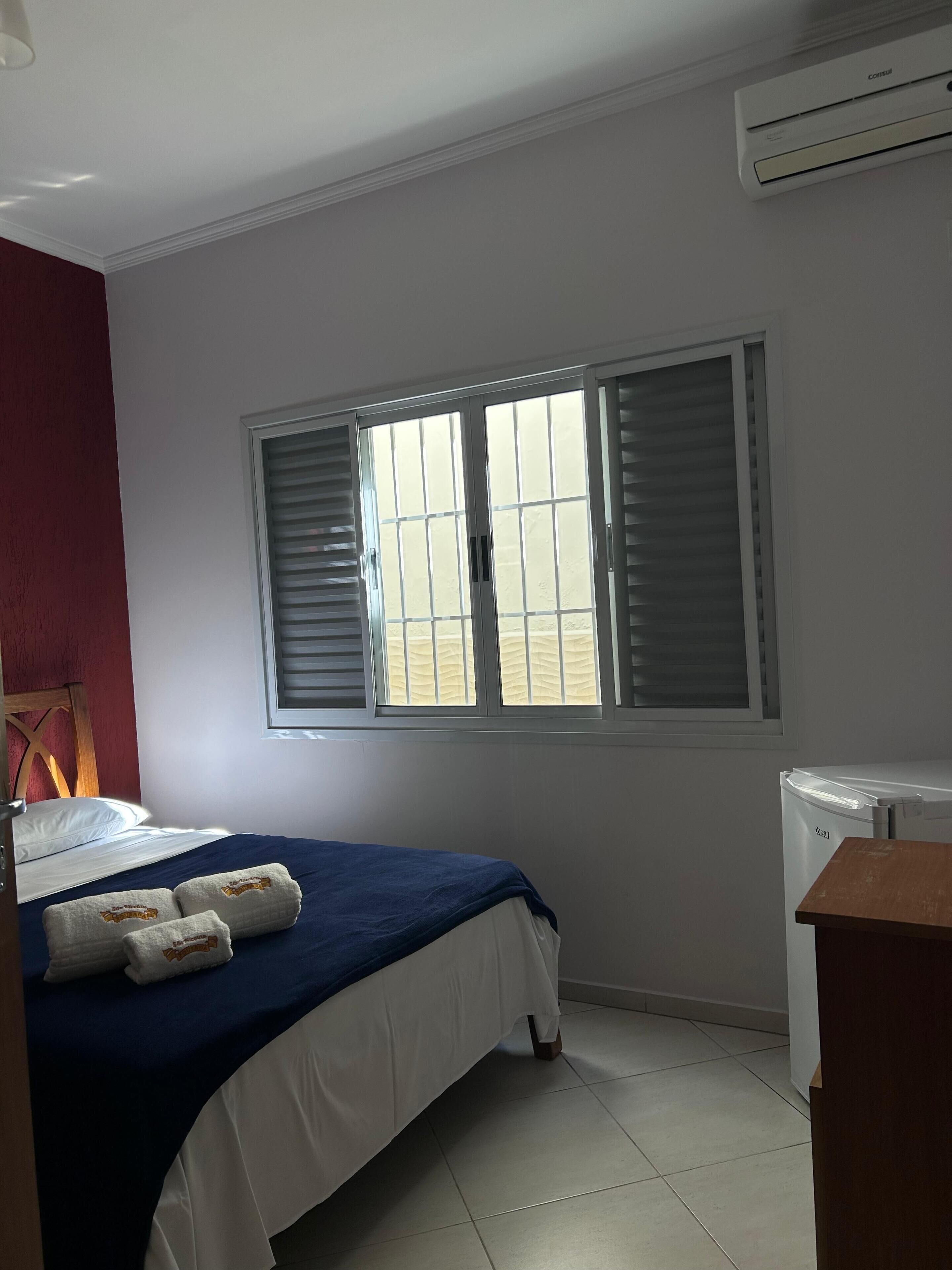 Quarto de hotel com cama, janela com portadas, ar condicionado branco e mesa de cabeceira de madeira.