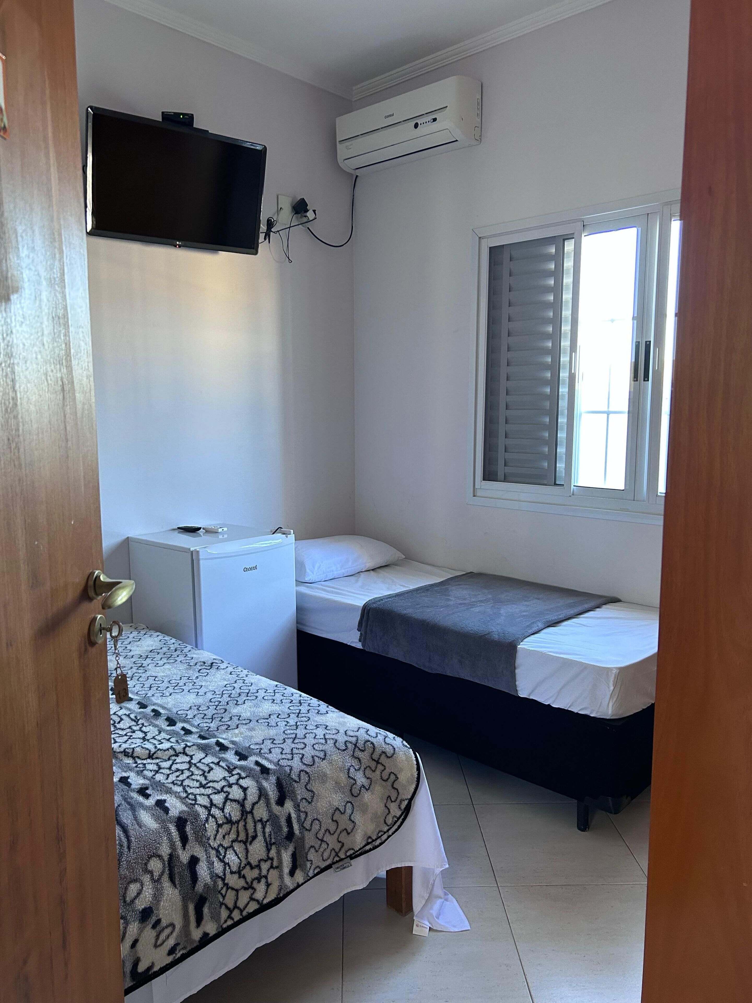 Quarto com duas camas, um ar condicionado branco, uma janela com persianas e uma porta com puxador de latão.