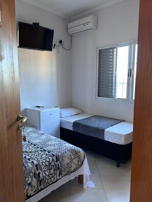 Quarto com duas camas, um ar condicionado branco, uma janela com persianas e uma porta com puxador de latão.