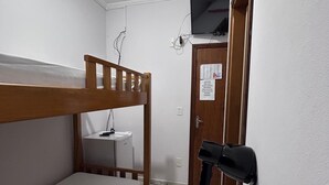 Quarto pequeno e compacto com beliche, escrivaninha com cadeira, secador de cabelo e televisão na parede.