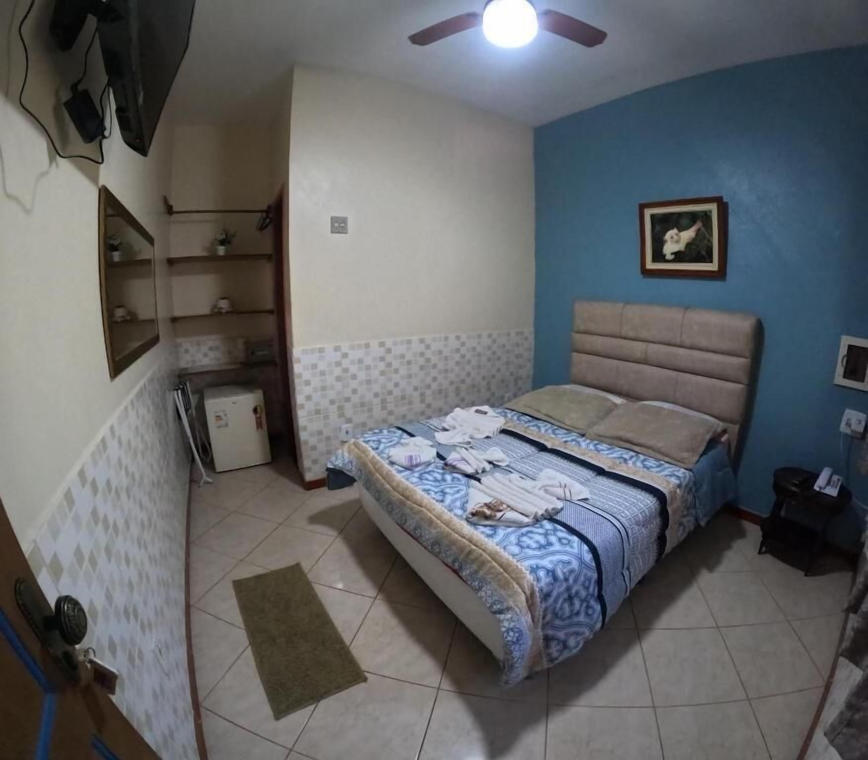 Quarto com cama, ventilador de teto, escrivaninha e televisão na parede.