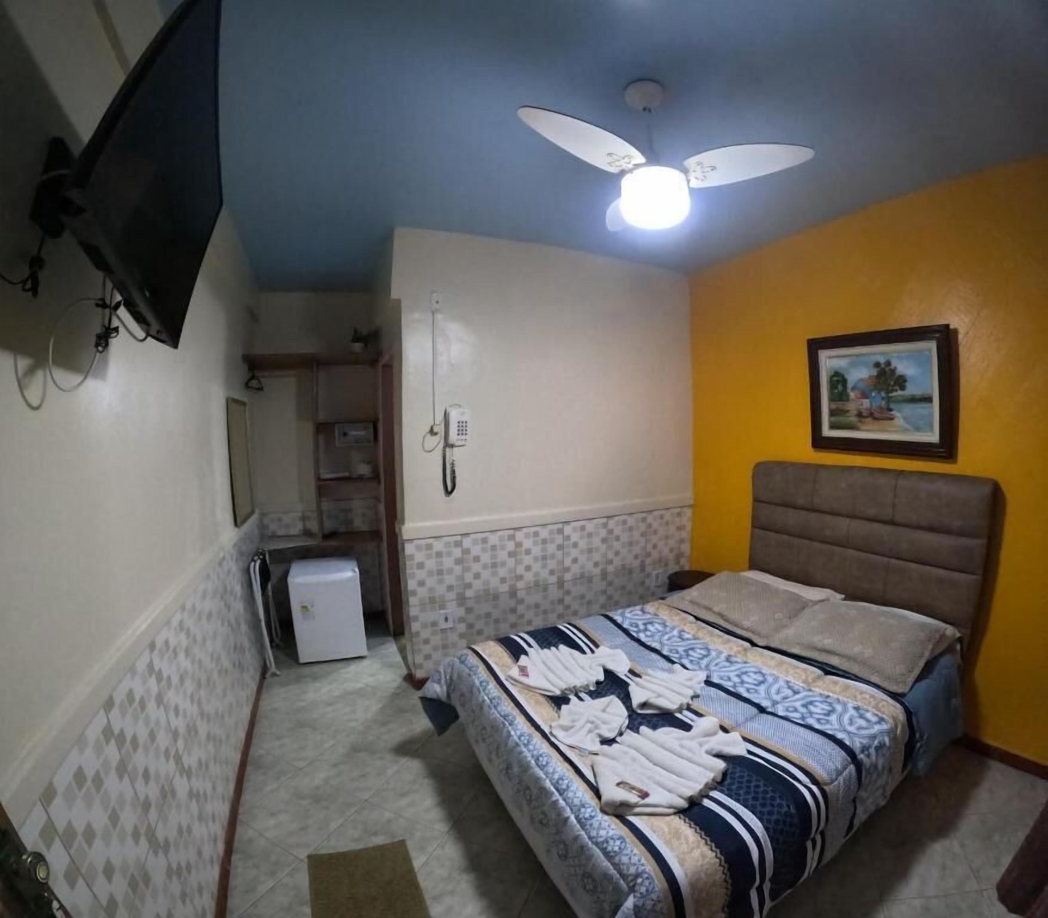 Quarto com cama, ventilador de teto, televisão fixada na parede e um pequeno frigorífico.