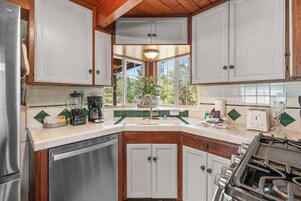 Uma cozinha com armários brancos, fogão, lava-louças e janela com vista para o verde.