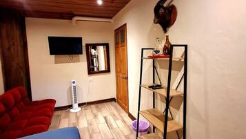 Quarto com cama, sofá vermelho, piso de madeira, ventilador de teto, televisão fixada na parede e prateleira com objeto decorativo.
