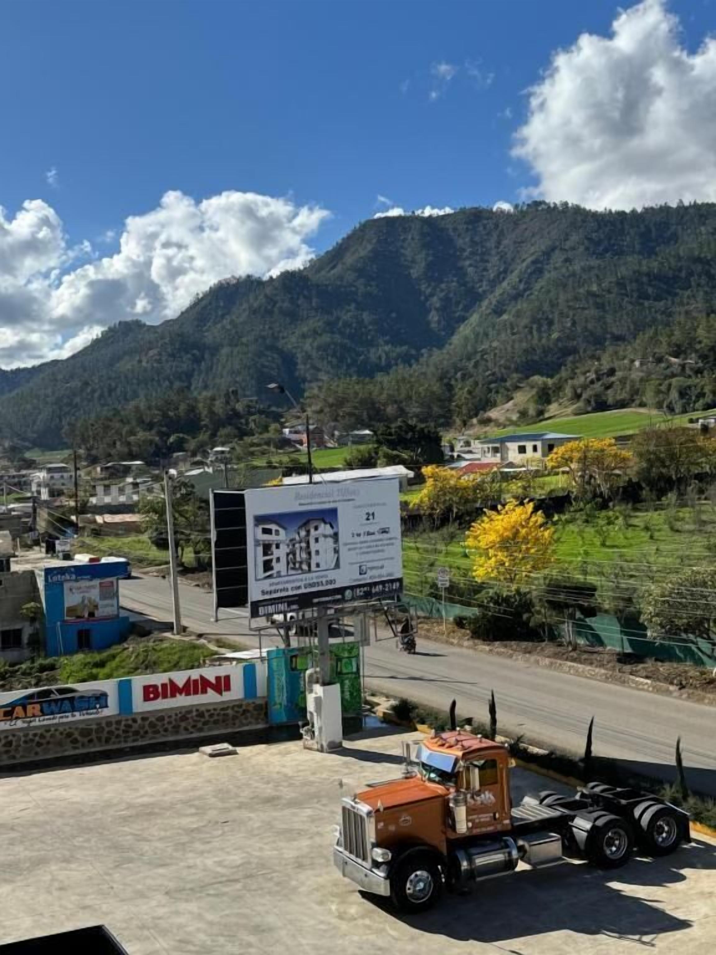 Um caminhão estacionado em área semiurbana, com montanhas ao fundo, um outdoor e um edifício com placa.