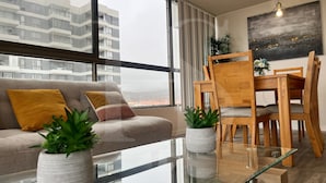 Uma sala de estar moderna com um sofá, duas mesas laterais com plantas, uma mesa de centro de vidro e um espaço de jantar com cadeiras e uma mesa.
