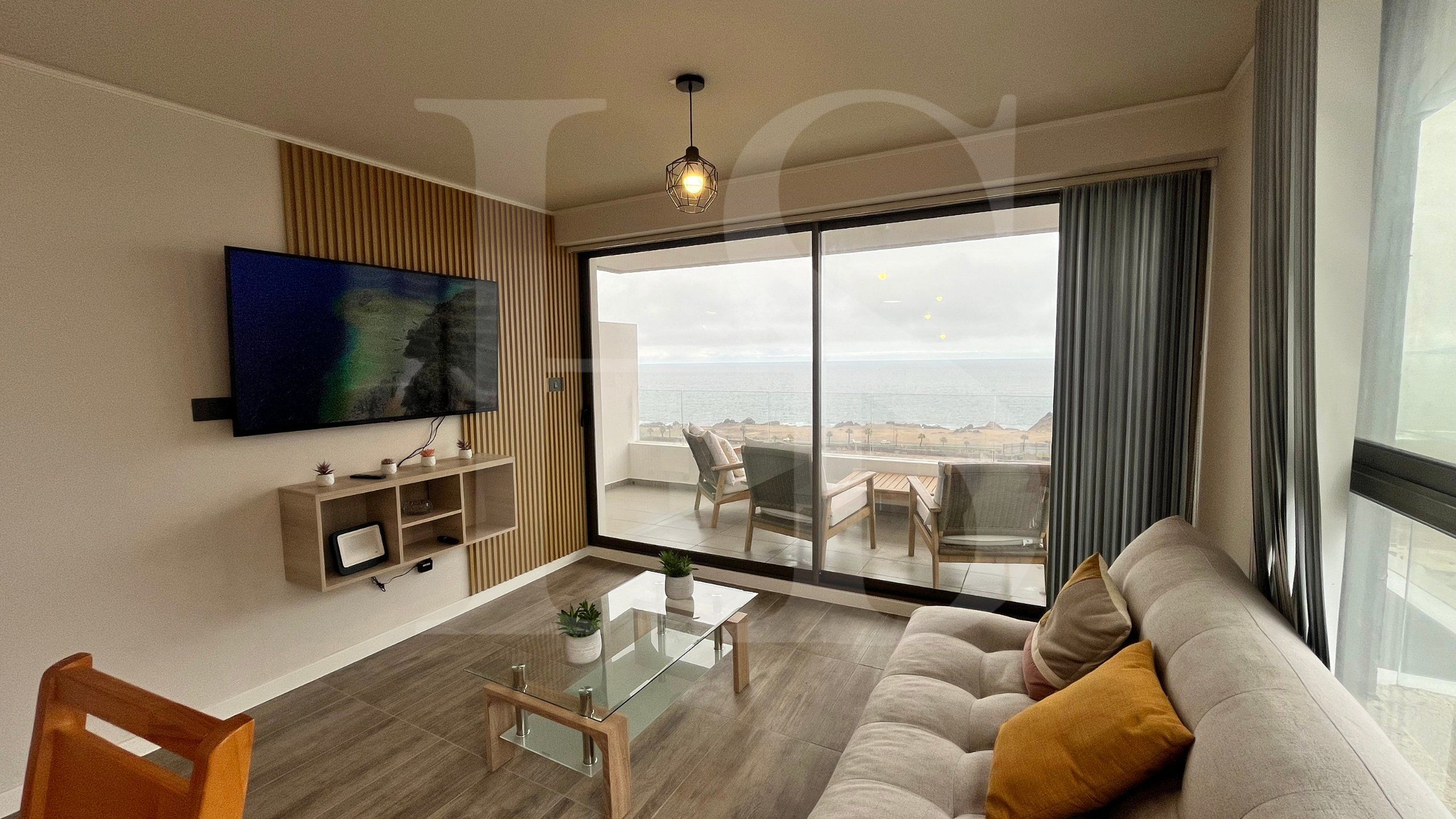 Uma sala de estar moderna com uma TV grande, uma mesa de centro de vidro, um sofá e vista para a praia.