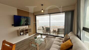 Uma sala de estar moderna com uma TV grande, uma mesa de centro de vidro, um sofá e vista para a praia.