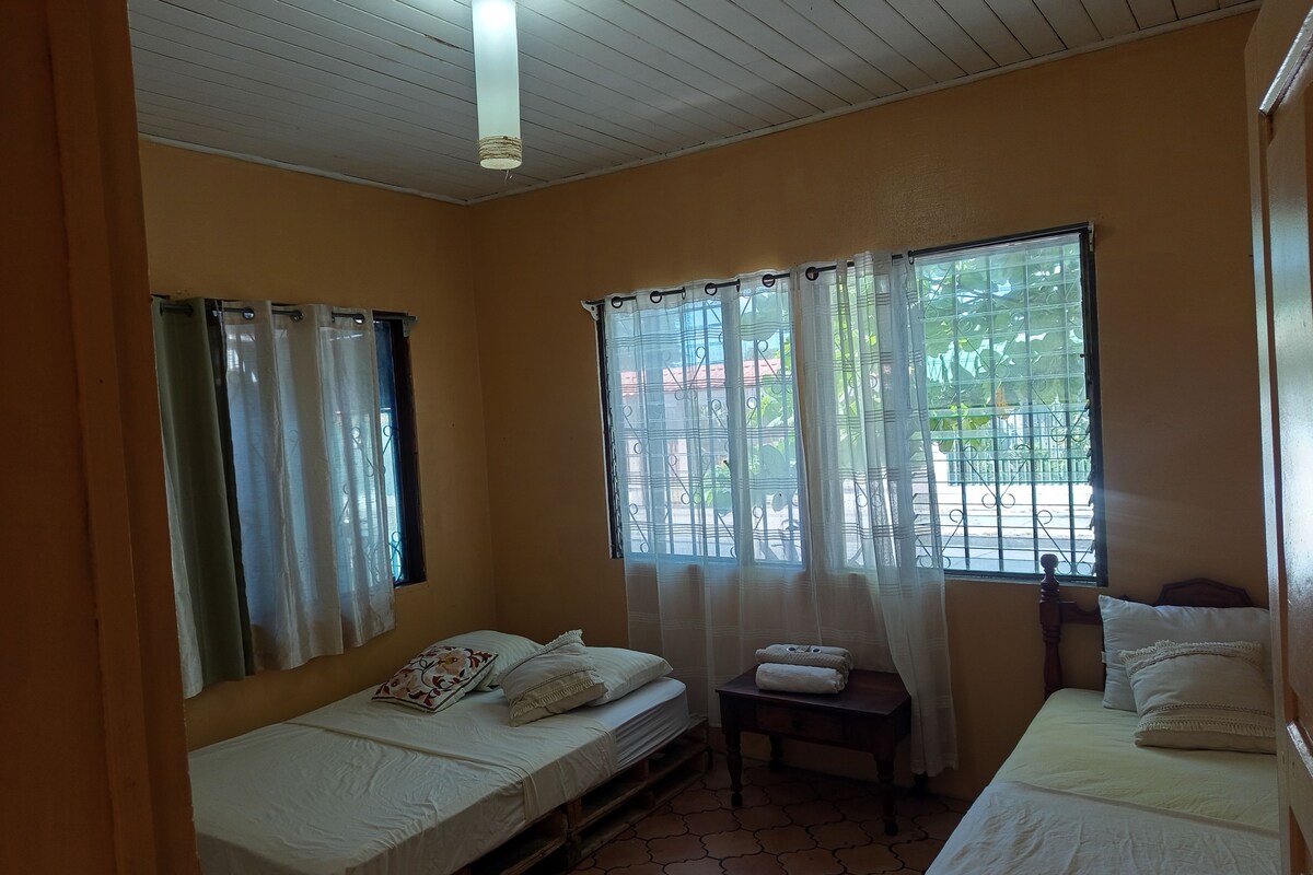 Quarto com duas camas, uma mesinha e uma janela com cortinas.
