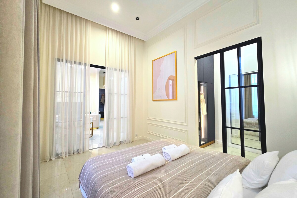 Quarto de hotel com cama, janela grande, porta e um quadro na parede.