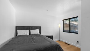 Quarto com cama, mesa de cabeceira, abajur e janela com persianas.