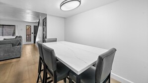 Uma sala de jantar moderna com uma mesa de mármore branco e cadeiras pretas, um sofá cinzento e soalho de madeira.