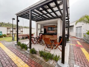 Um pátio externo com pérgola, mesa de jantar com cadeiras, área de cozinha com televisão e um caminho acessível para cadeiras de rodas.