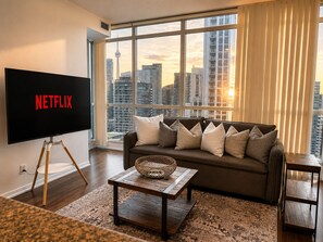 Uma sala de estar moderna com uma televisão grande exibindo 'NETFLIX', um sofá marrom com várias almofadas, uma mesa de centro de madeira e vista para um horizonte urbano com uma torre.