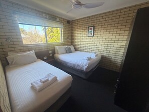 Quarto de hotel com duas camas, janela com persianas, ventilador de teto e parede de tijolos.