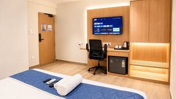 Quarto de hotel com cama, escrivaninha com computador, cadeira, pequeno frigorífico e televisão na parede.