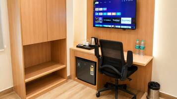 Quarto de hotel com armário de madeira, uma escrivaninha com monitor de computador, uma cadeira de escritório preta, uma cafeteira e um cesto de lixo.