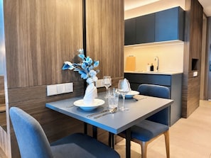 Uma área de jantar moderna com uma mesa posta para dois, um vaso com flores azuis e uma cozinha ao fundo.