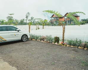 Um carro branco com a marca 'THE OKA' estacionado em frente a um hotel com um recurso de água e plantas tropicais.