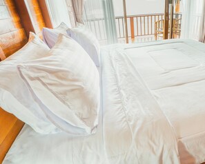 Uma cama com roupa de cama branca, cabeceira de madeira e vista para uma varanda com guarda-corpo.