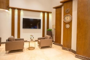 Um lobby moderno com televisão de tela plana, dois sofás, uma mesa redonda pequena com um vaso e um pilar de madeira com relógios.