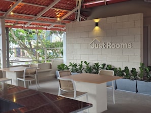 Espaço de café com placa 'JustRooms', mesas ao ar livre e plantas em vasos.
