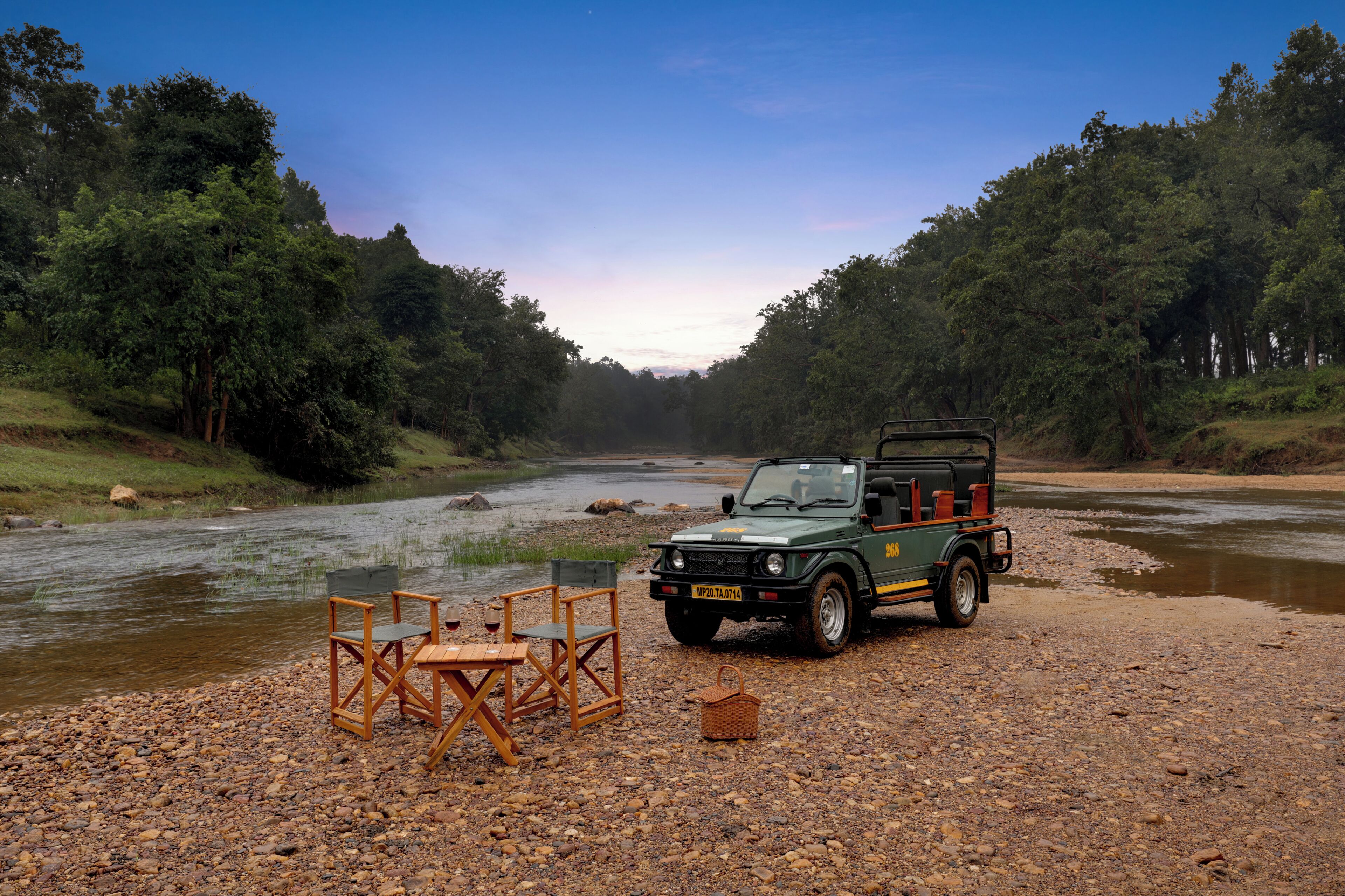 Veículo safari verde estacionado em área de cascalho, com um rio ao fundo e floresta de ambos os lados.