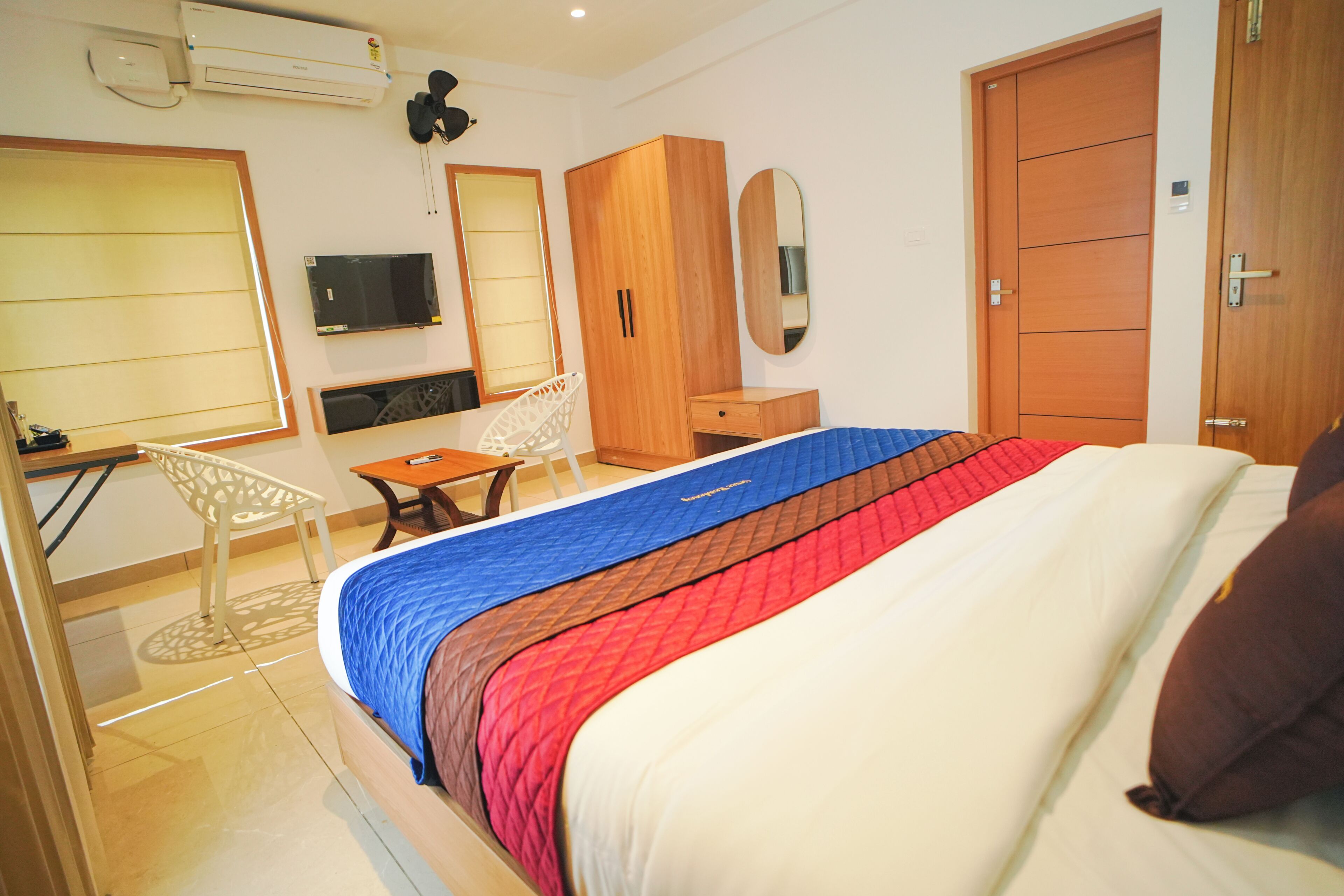 Quarto de hotel com cama, secretária, cadeira, guarda-roupa, televisão e janela.