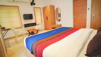 Quarto de hotel com cama, secretária, cadeira, guarda-roupa, televisão e janela.