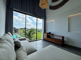 Uma sala de estar moderna com uma janela grande, um sofá, um móvel de TV de madeira e um lustre.