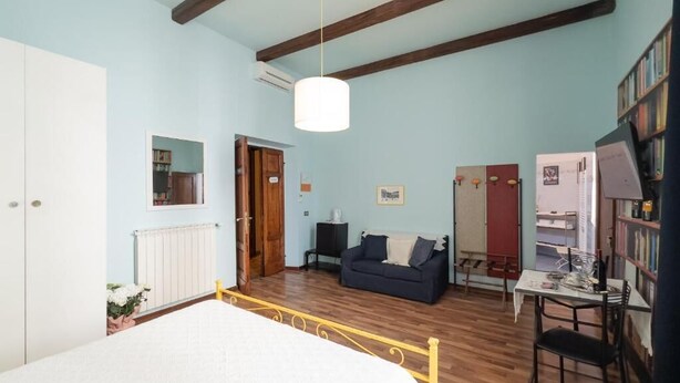 Quarto com cama, piso de madeira, radiador branco, parede azul, teto de madeira, porta branca, guarda-roupa de madeira, mesa de cabeceira branca, luminária branca, sofá azul, mesa de madeira, uma cadeira e estante.