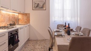 Uma cozinha compacta com fogão a gás, forno e pia, uma mesa de jantar para dois com cadeiras, e uma janela com cortinas leves.