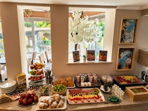 Buffet com diversos alimentos, incluindo frutas, doces e queijos, disposto em um balcão com vista para o exterior.