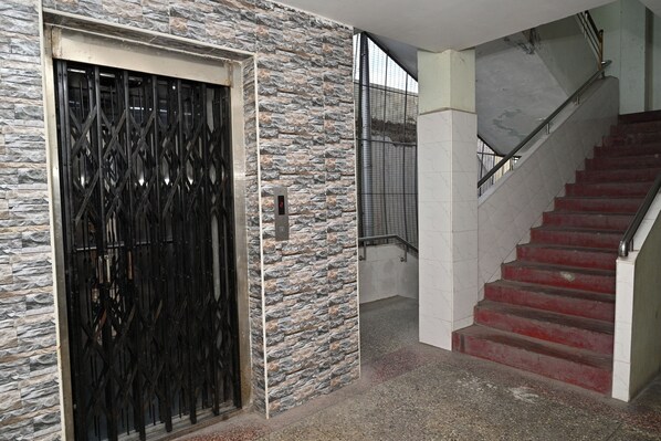 Uma escadaria com degraus vermelhos que levam a um andar superior, um portão de metal com visor digital e uma parede de pedra com textura.