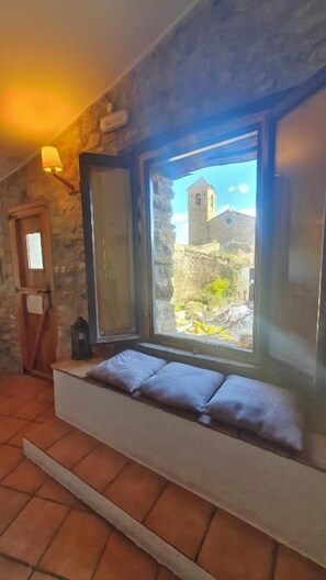 Quarto com parede de pedra, janela com vista para um edifício e um banco com almofadas.