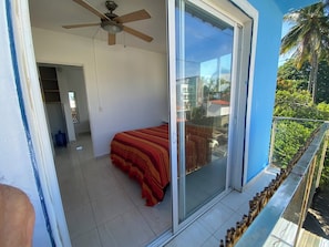 Quarto com cama, ventilador de teto e porta de vidro deslizante que dá para área externa com vista para vegetação e rua.