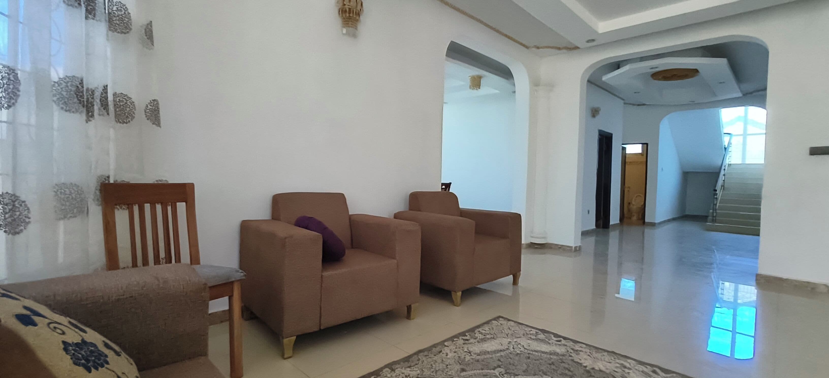 Uma sala ampla com teto alto, uma cadeira de madeira, dois poltronas e um tapete estampado.