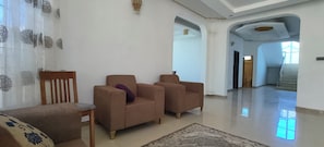 Uma sala ampla com teto alto, uma cadeira de madeira, dois poltronas e um tapete estampado.