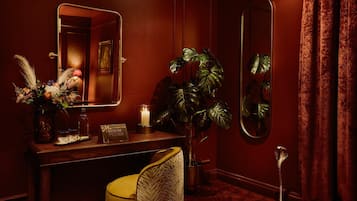 Um quarto com iluminação suave, uma mesa consola de madeira, um espelho, um vaso com planta, uma vela e uma cadeira.