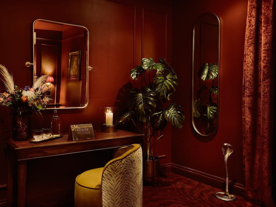 Um quarto com iluminação suave, uma mesa consola de madeira, um espelho, um vaso com planta, uma vela e uma cadeira.