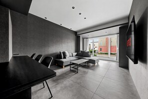 Uma sala de estar moderna com sofá, mesa de centro e cadeiras, uma TV de tela plana na parede e uma janela grande com vista para o exterior.
