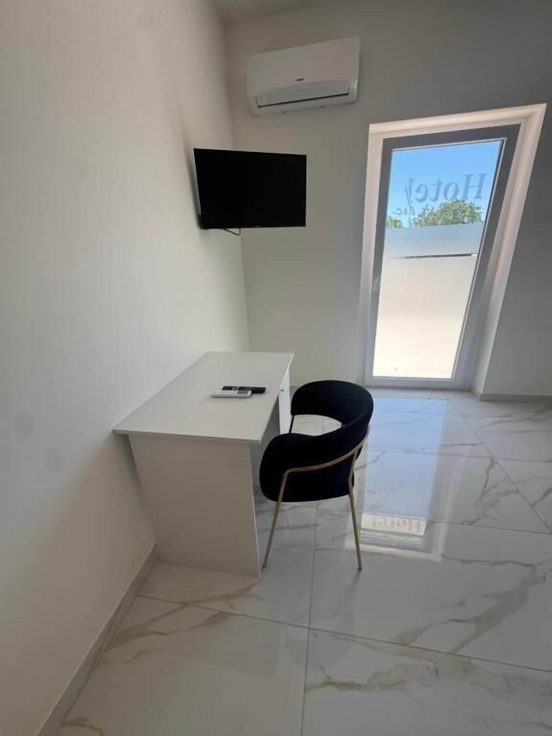 Quarto com escrivaninha branca, cadeira preta, televisão de tela plana na parede e porta de vidro deslizante para área externa.