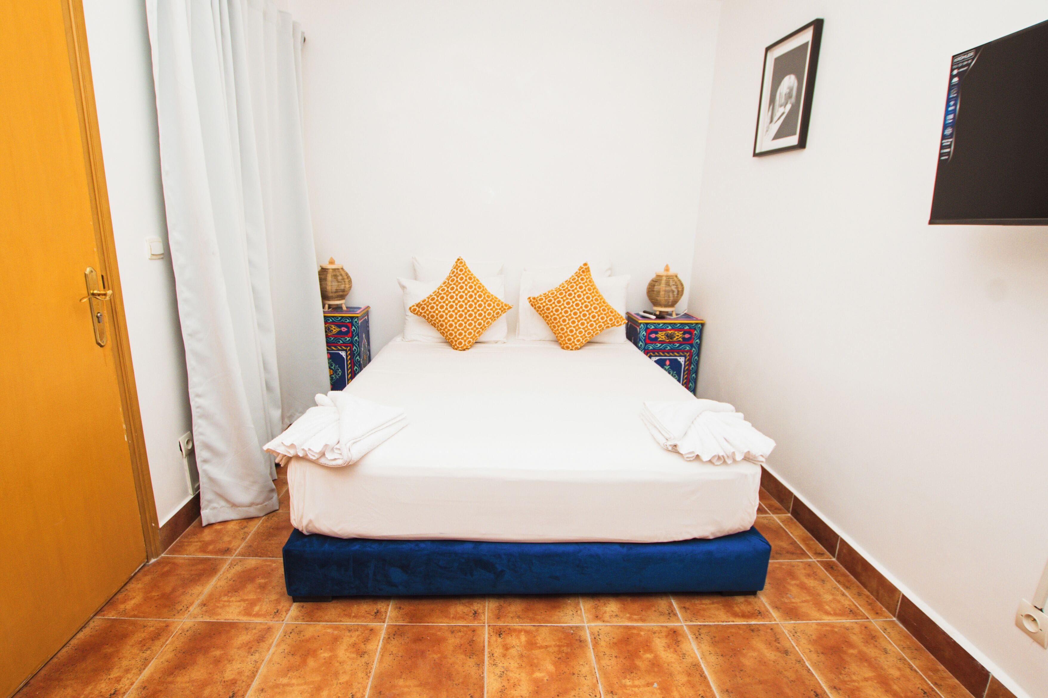 Quarto de hotel com cama, almofadas, mesa de cabeceira, mesinha e um quadro na parede.