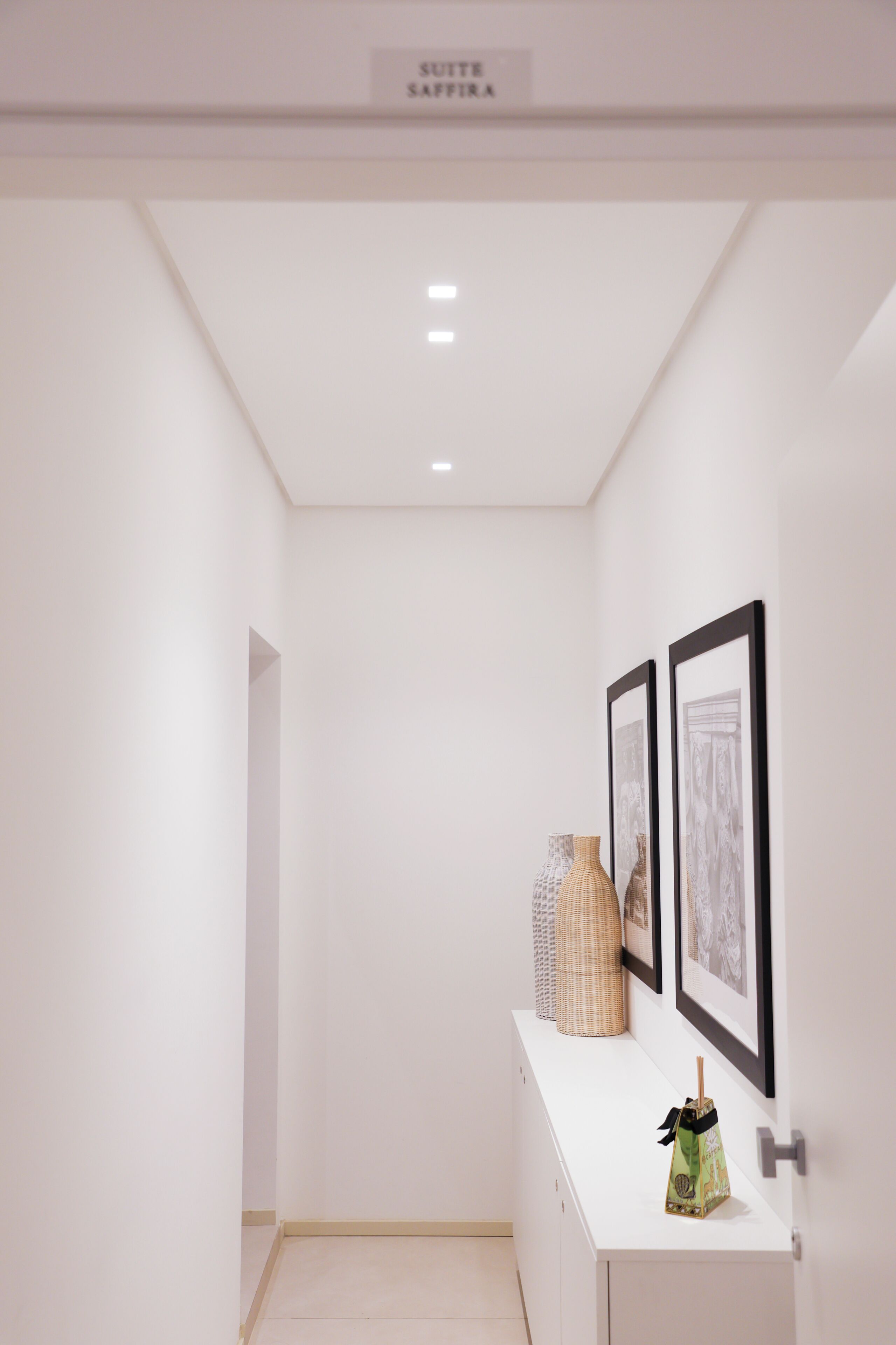 Um corredor com parede branca, teto com iluminação embutida e armário branco com duas molduras e um vaso decorativo.